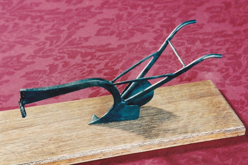 Dark-coloured metal plough set on pine-like timber plank. ; 1995; 19894 ...